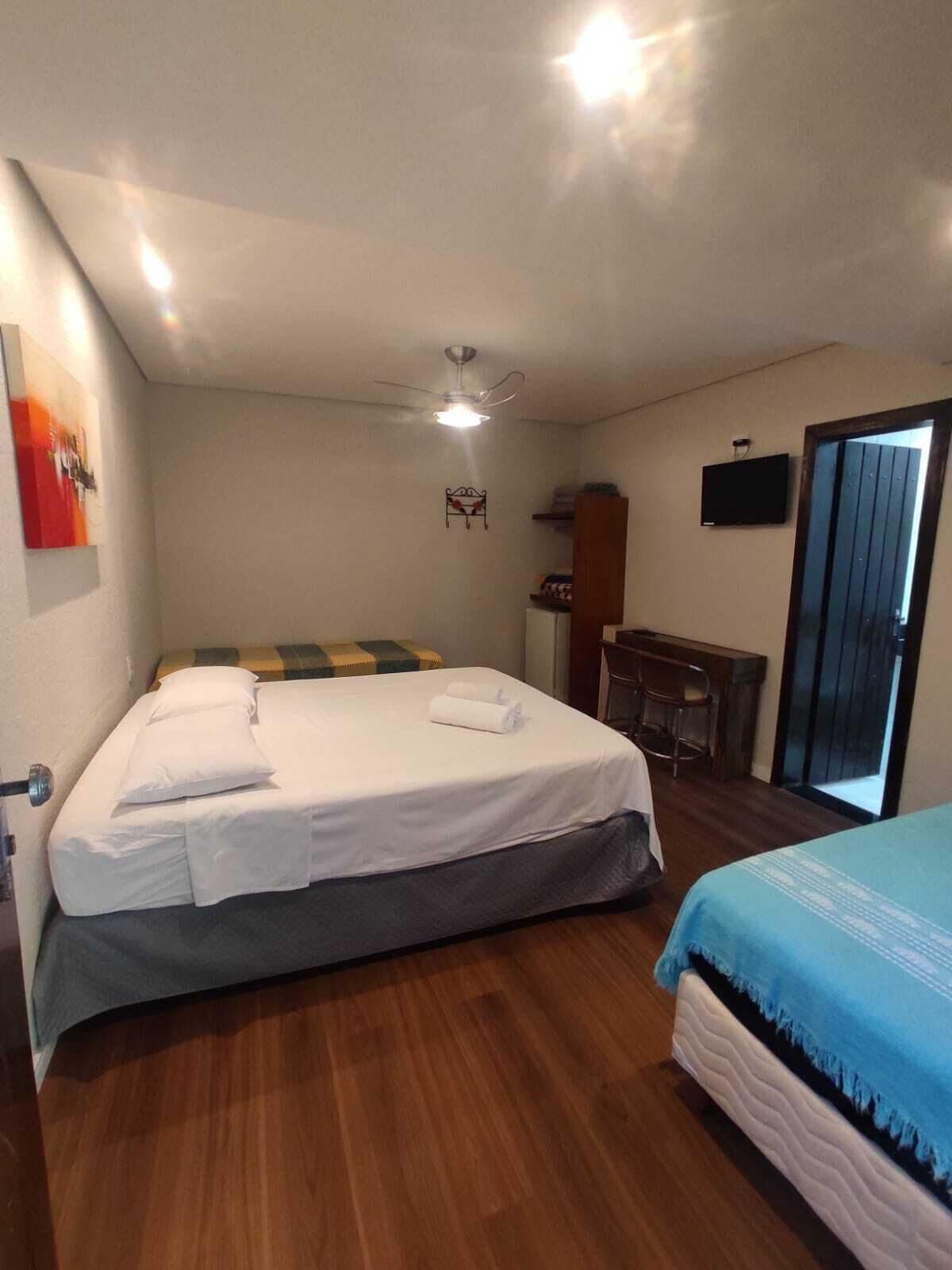 Quarto com cama, escrivaninha, cadeira, ventoinha no teto e televisão.