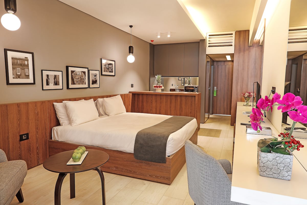 Quarto de hotel moderno com uma cama grande, uma mesinha, uma cadeira e um espaço com cozinha.