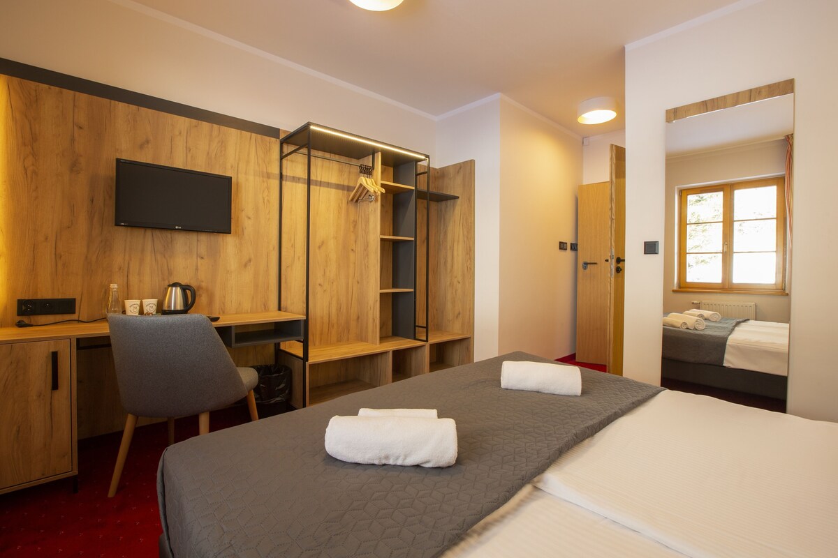 Quarto de hotel com cama, escrivaninha, cadeira, televisão, guarda-roupa e janela.