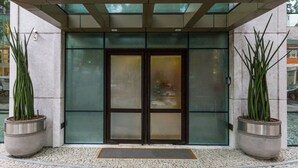 Entrada de um prédio com porta de vidro, duas plantas em vasos e piso revestido com azulejos.