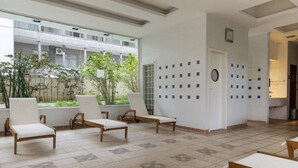 Um quarto com piso de azulejos, várias cadeiras de madeira com almofadas, uma janela grande e uma parede com um padrão de azulejos quadrados.