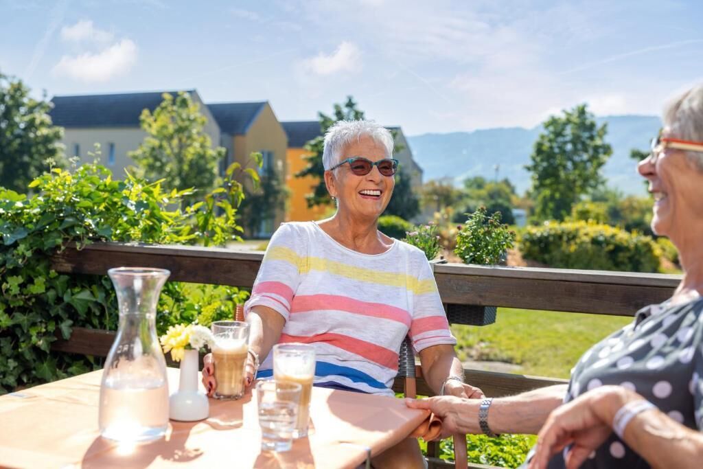 Duas pessoas sentadas em uma mesa ao ar livre, com bebidas e um vaso de flores, com vista para o verde e prédios.