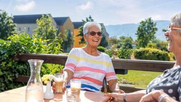 Duas pessoas sentadas em uma mesa ao ar livre, com bebidas e um vaso de flores, com vista para o verde e prédios.