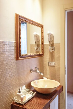 Um banheiro com parede revestida de azulejos bege, um espelho grande com moldura de madeira, uma pia com cuba bege e dois secadores de cabelo fixados na parede.