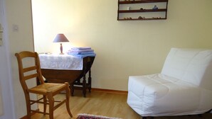 Um quarto com uma cadeira de madeira, uma mesa pequena com abajur, cama branca e tapete estampado.