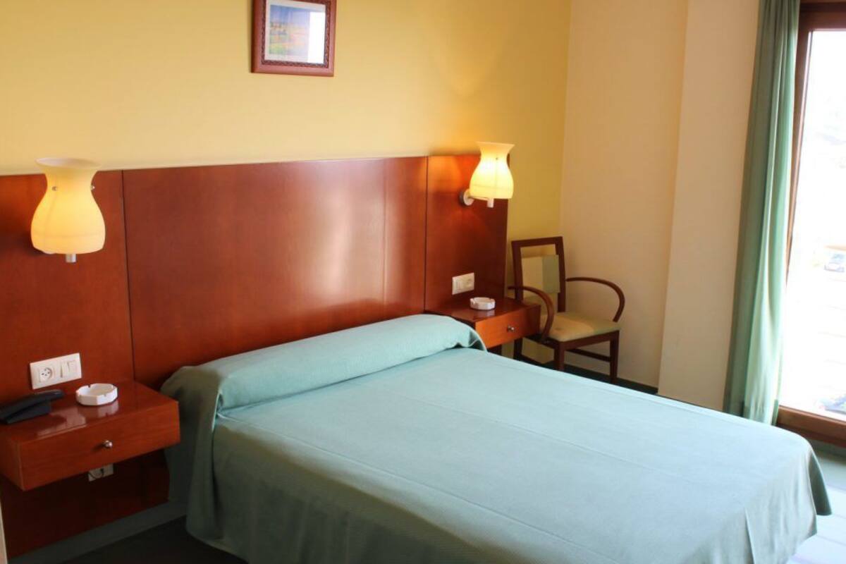 Quarto de hotel com cama, luminárias de cabeceira, uma cadeira, janela e uma mesinha com controle remoto.