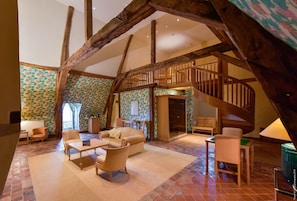 Um amplo loft com vigas de madeira expostas, uma área de estar aconchegante com sofá e poltronas, uma mesa de jantar e uma escada que leva a um andar superior.