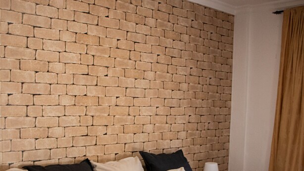Quarto de hotel com cabeceira em padrão de tijolos, cama com várias almofadas, mesa de cabeceira com abajur e janela com cortinas.