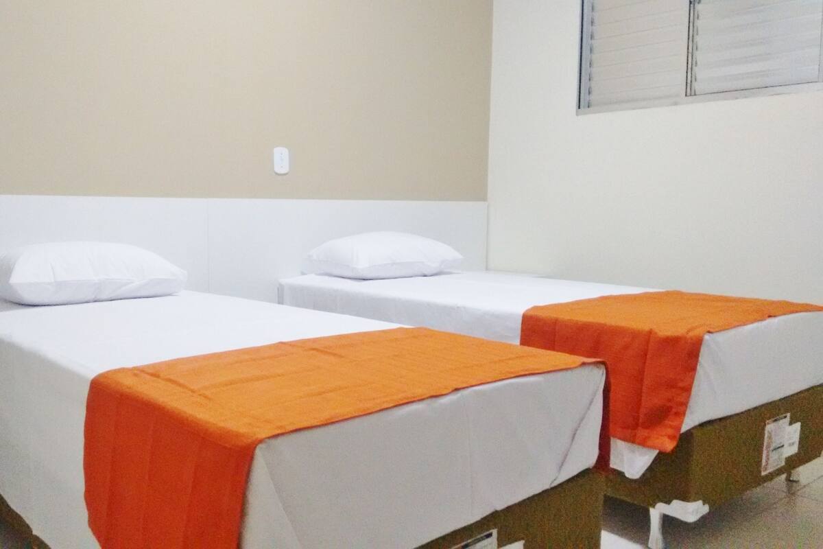 Quarto de hotel com duas camas de solteiro, cada uma com roupa de cama branca e manta laranja. Há uma cadeira no canto e uma janela com persianas na parede.