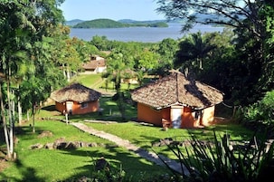 Um resort tropical com chalés de telhado de palha, vegetação exuberante e vista para um lago.