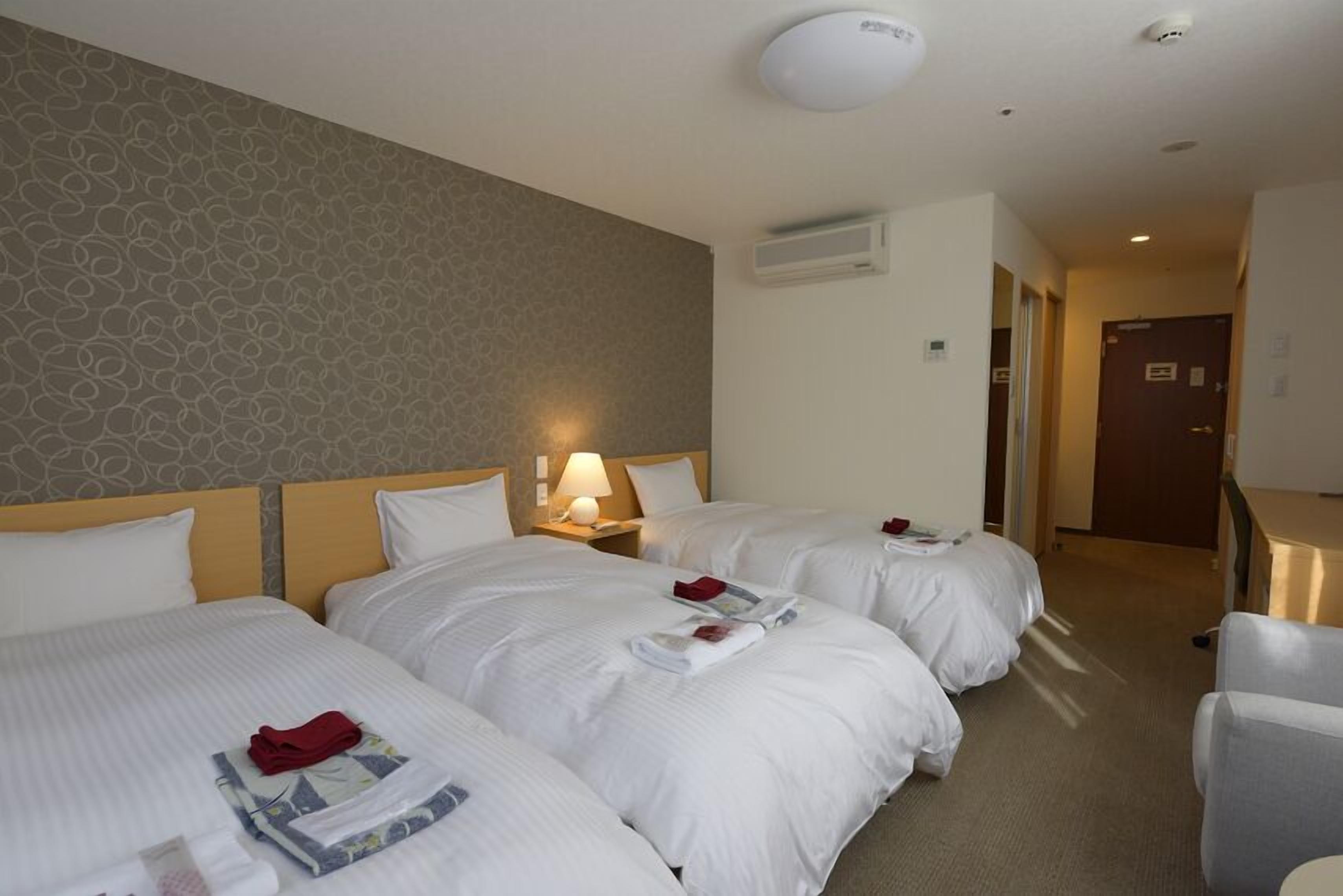 Quarto de hotel com três camas, uma escrivaninha, uma cadeira e uma porta.