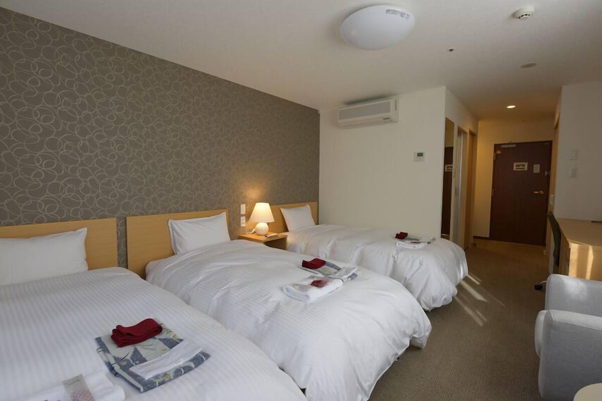 Quarto de hotel com três camas, uma escrivaninha, uma cadeira e uma porta.