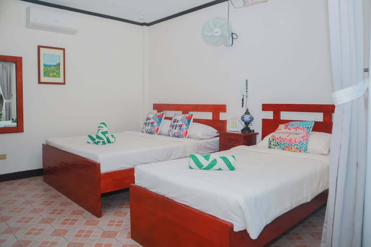 Quarto de hotel com duas camas de madeira, um ventilador de teto, uma mesa de cabeceira e uma janela com cortinas.