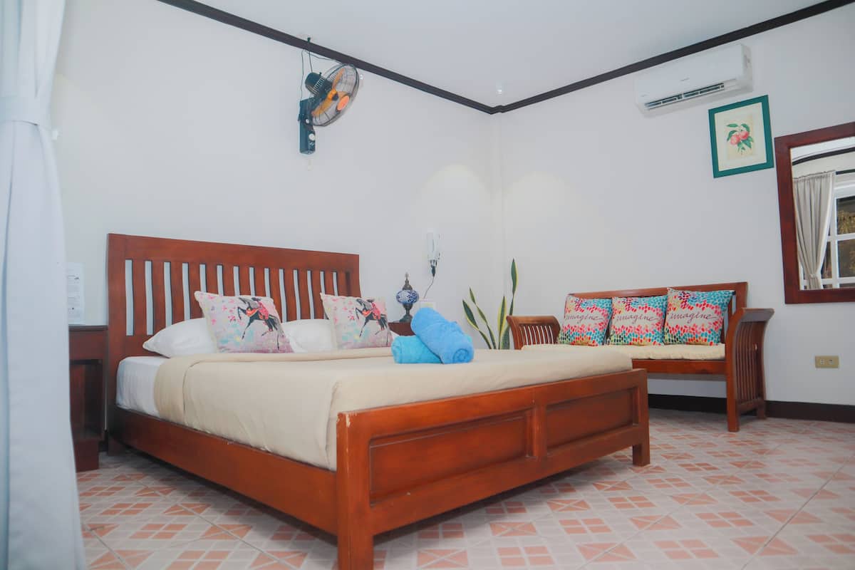 Quarto com cama de madeira, mesa de cabeceira, cadeira, ventoinha e janela com cortinas.