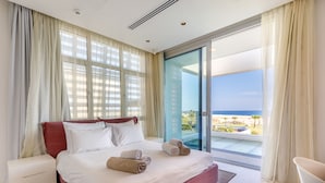 Quarto de hotel com uma cama grande, mesa de cabeceira e porta de vidro que dá para uma varanda com vista para a praia.