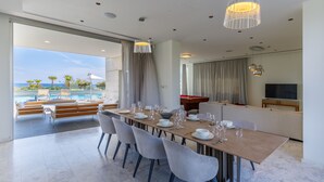 Uma área de jantar moderna com uma mesa comprida posta para seis pessoas, piscina visível pelas portas de vidro e um espaço de estar à beira da piscina.
