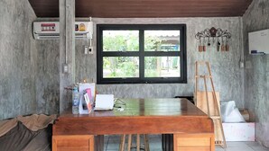 Quarto com mesa de madeira, janela com vista para vegetação, quadro branco e ar condicionado fixado na parede.