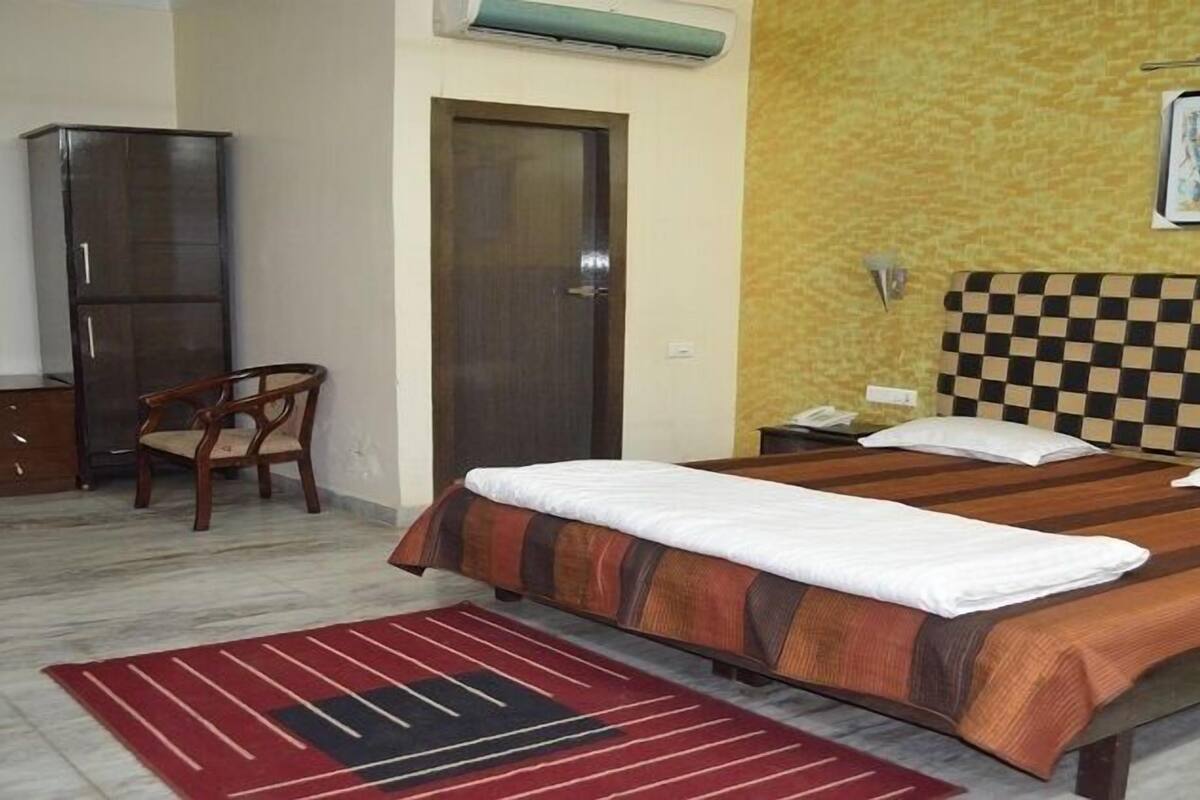 Quarto com cabeceira estampada em xadrez, cama com duas almofadas, mesa de cabeceira, cadeira, guarda-roupa e tapete listrado em vermelho e preto.