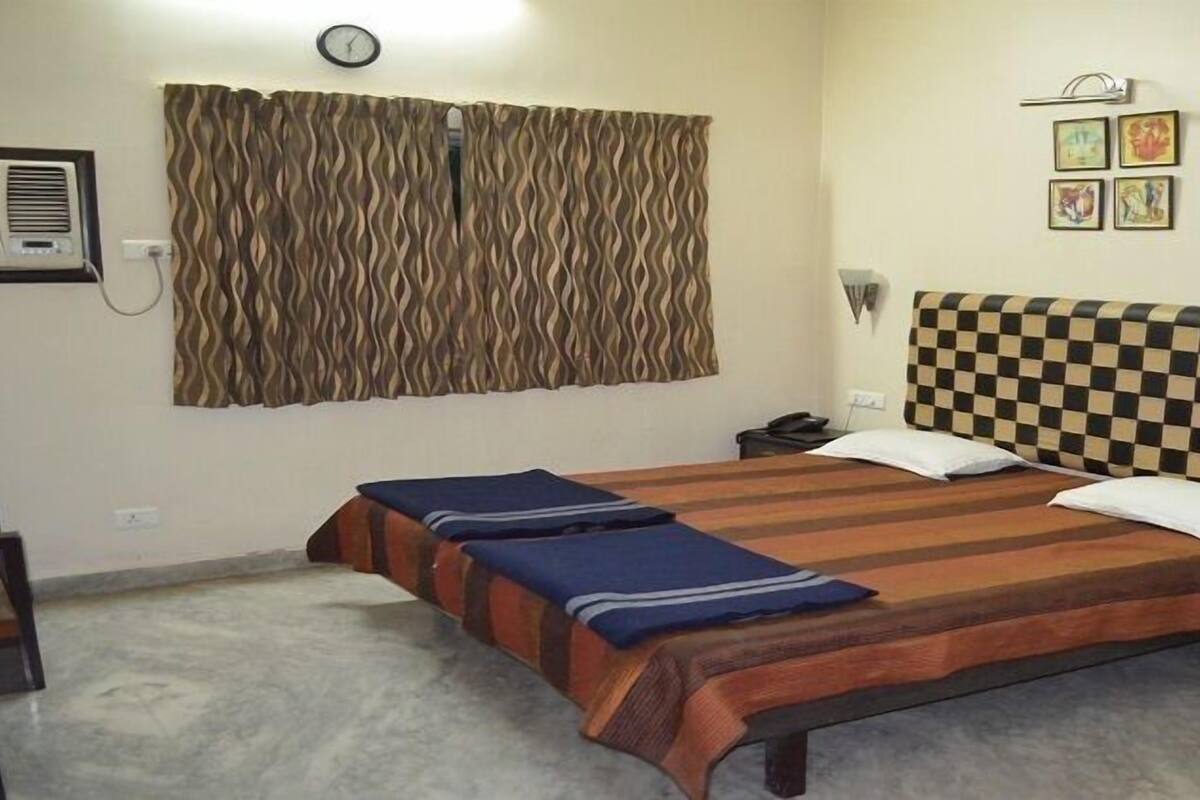 Quarto com cabeceira xadrez, cama com roupa de cama listrada, mesa de cabeceira, relógio de parede e janela com cortinas estampadas.