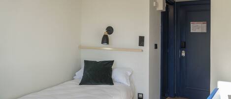 Quarto com cama, luminária de cabeceira, prateleira na parede, porta com placa e interruptor de luz.
