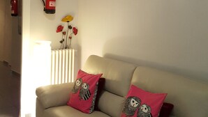 Uma sala de estar com um sofá bege, duas almofadas vermelhas com estampas de corujas, uma silhueta de touro preto, um extintor de incêndio, um radiador e uma pintura com vista para a cidade.