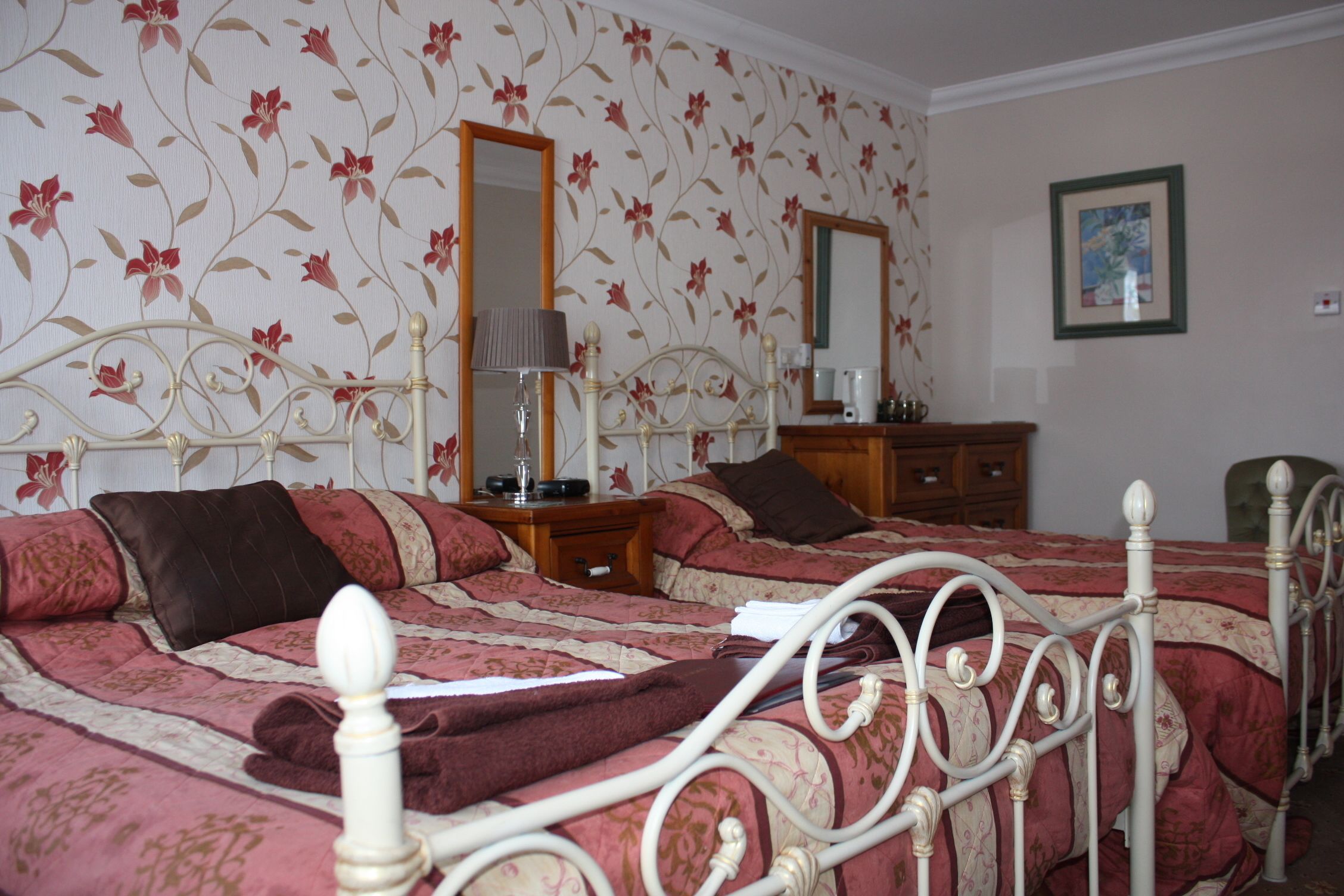 Quarto com papel de parede floral, cama com estrutura de metal, mesa de cabeceira com abajur e cômoda.