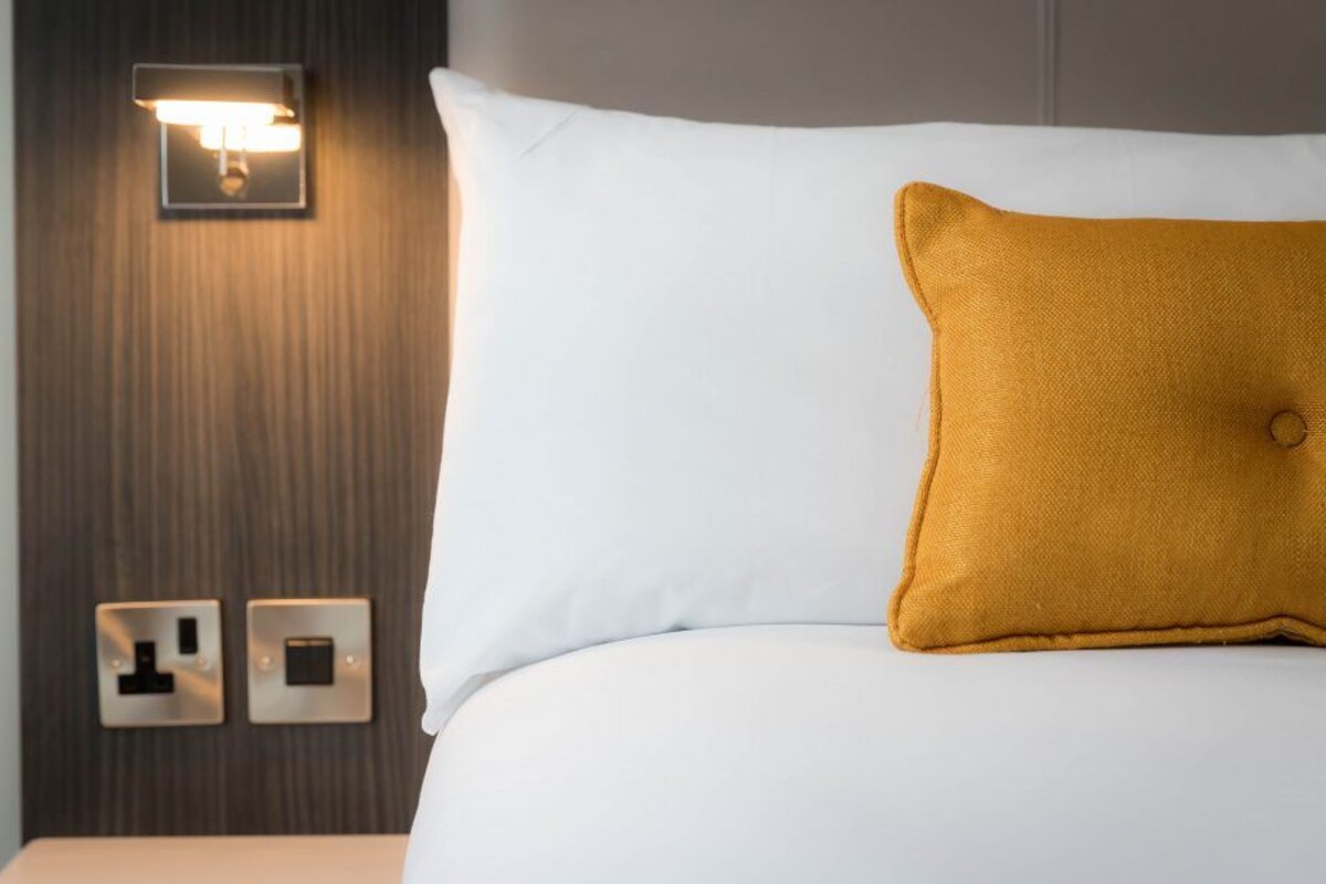 Quarto de hotel com uma cama, luminária fixada na parede e duas tomadas elétricas.