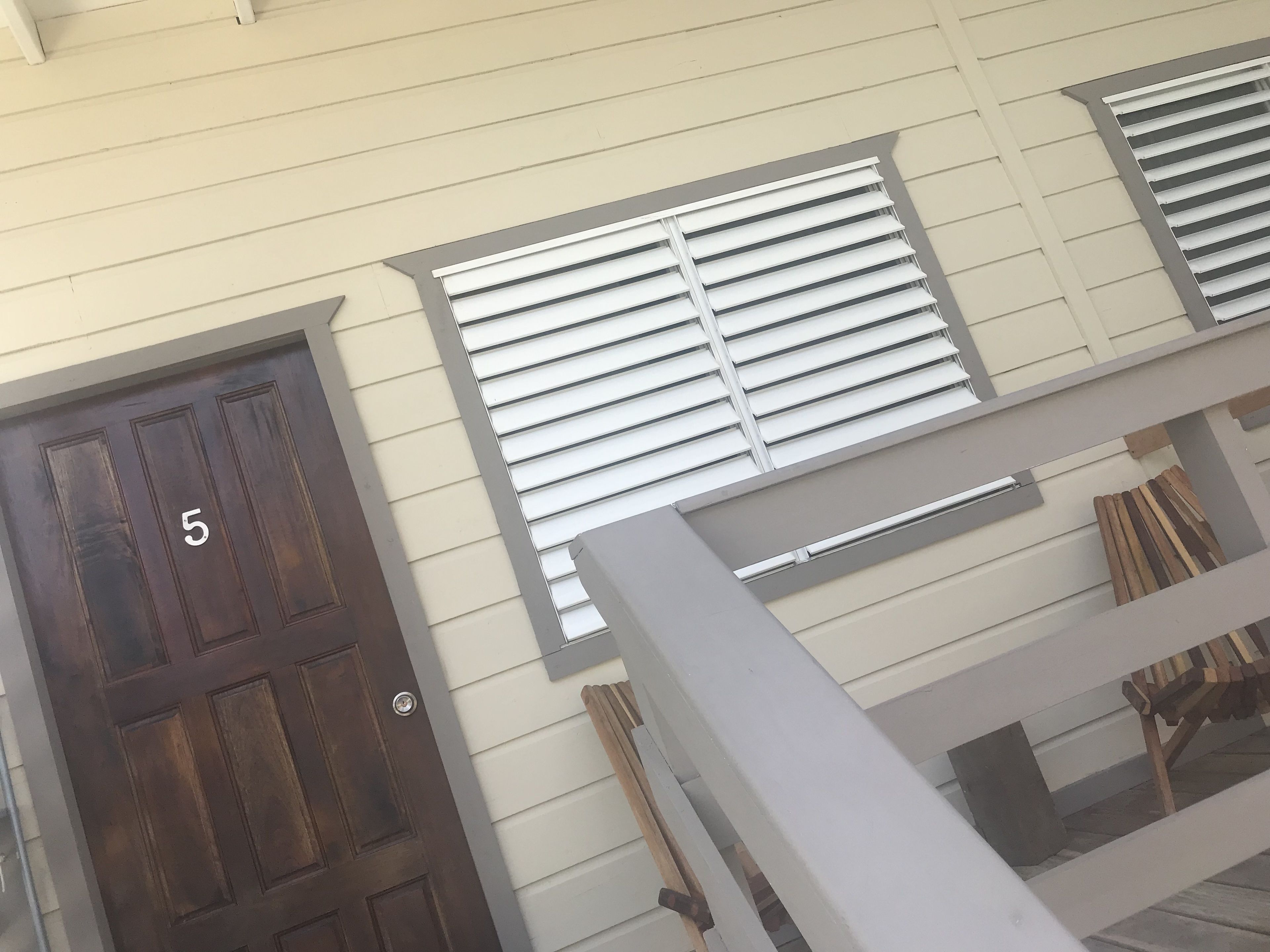 Uma casa com porta de madeira escura marcada com o número 5, revestimento branco e janela com persianas horizontais.