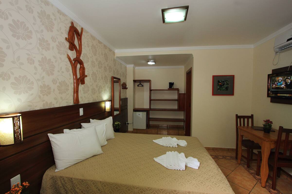 Quarto de hotel com cama, escrivaninha, cadeira, luminária, televisão e espelho.