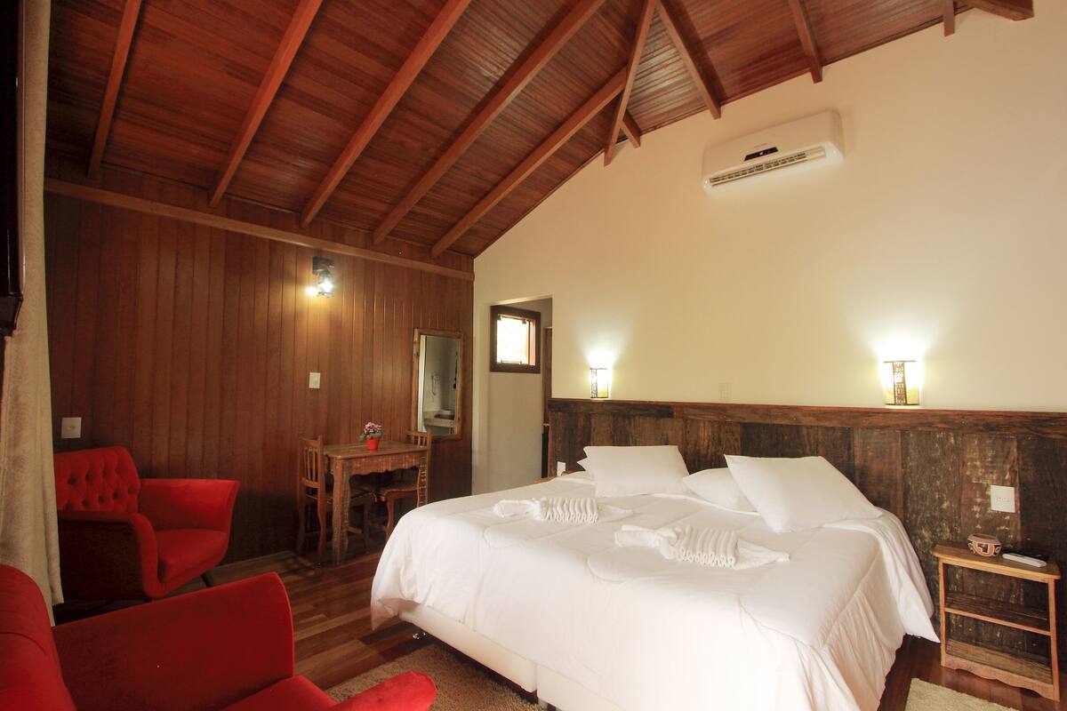 Quarto com teto de madeira, cama com roupa branca, poltrona vermelha, escrivaninha de madeira e um espelho.