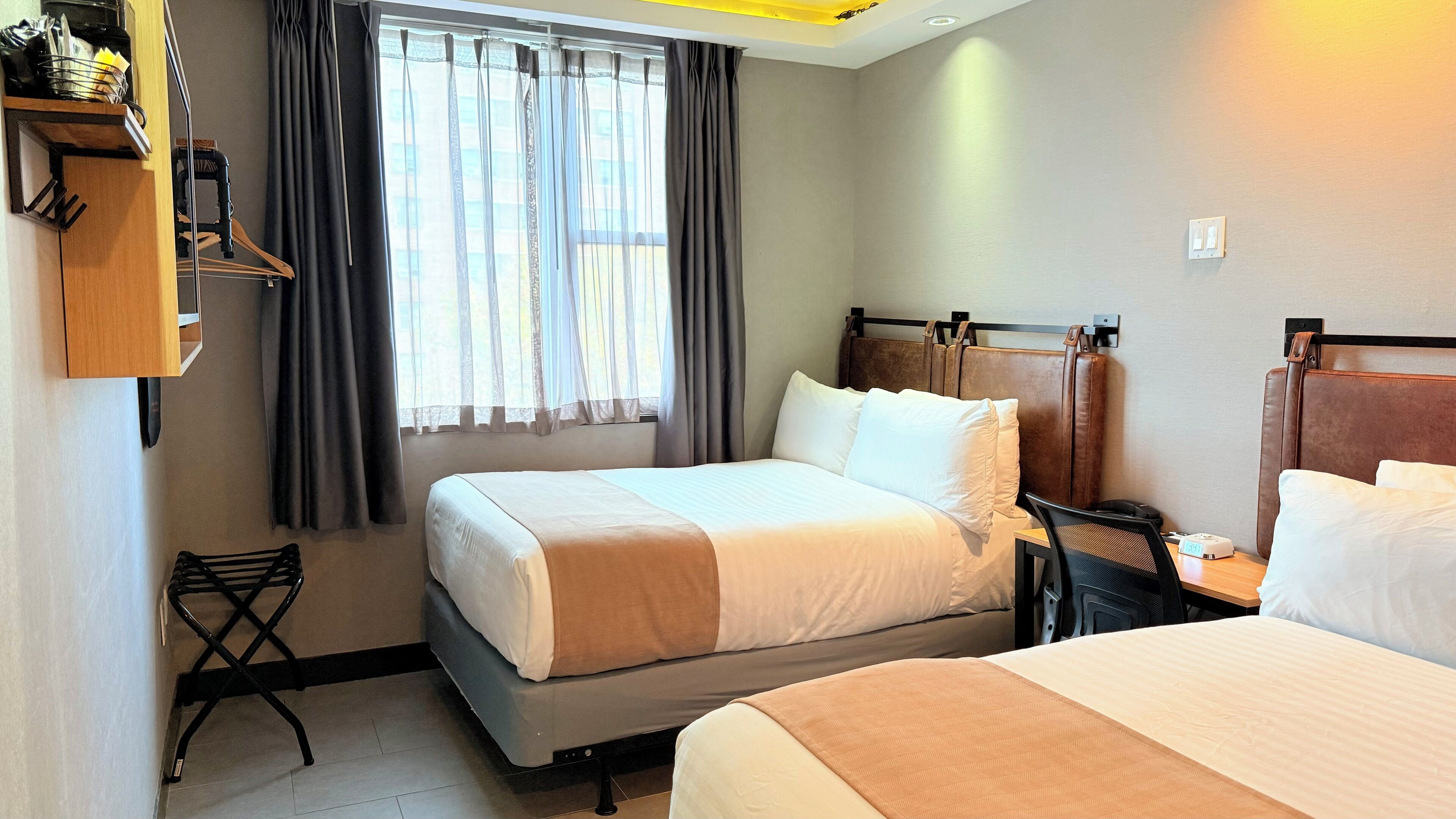 Quarto de hotel com duas camas, mesa de cabeceira, uma cadeira, prateleira com artigos de higiene pessoal e janela com cortinas.