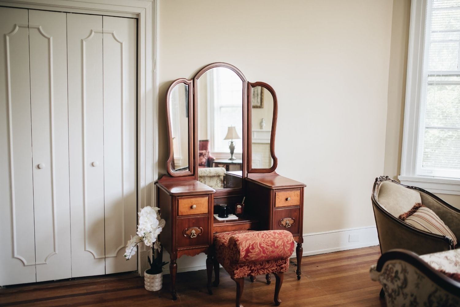 Quarto com toucador de madeira, espelho, um banquinho pequeno e uma cadeira.