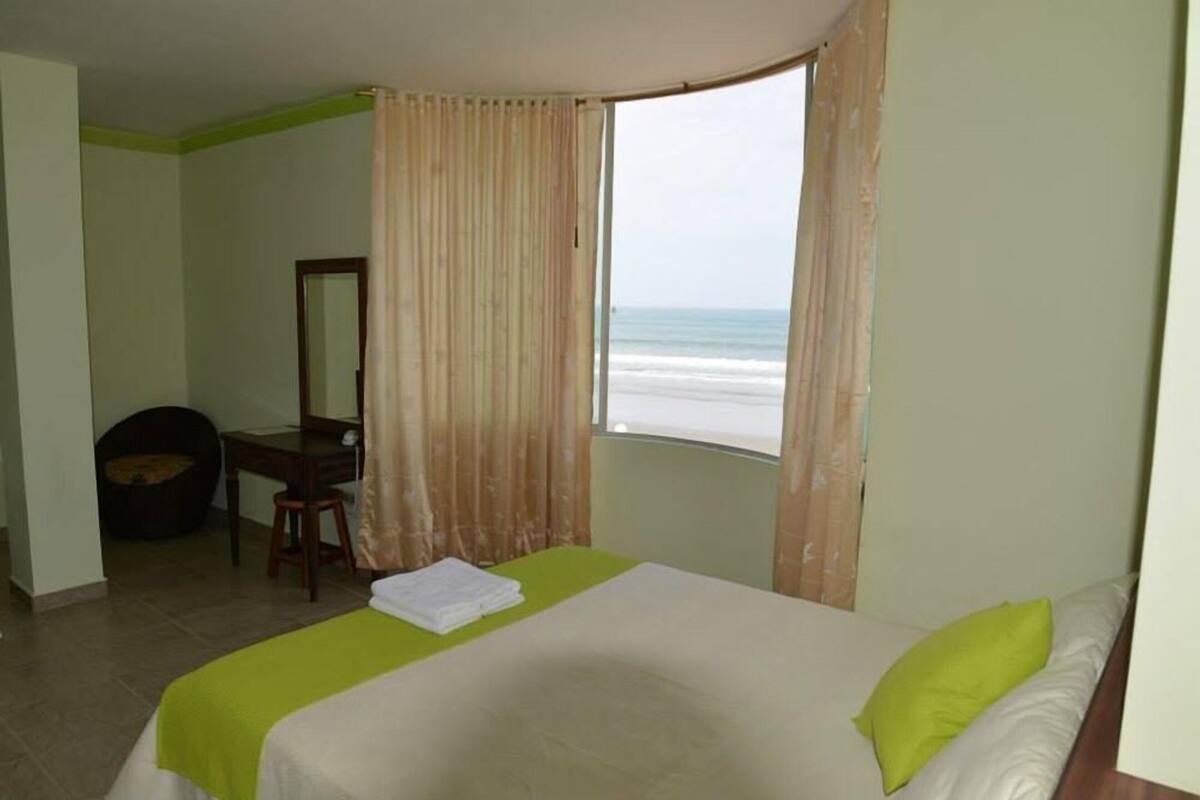 Quarto com cama, cadeira, escrivaninha, espelho e janela com vista para a praia.