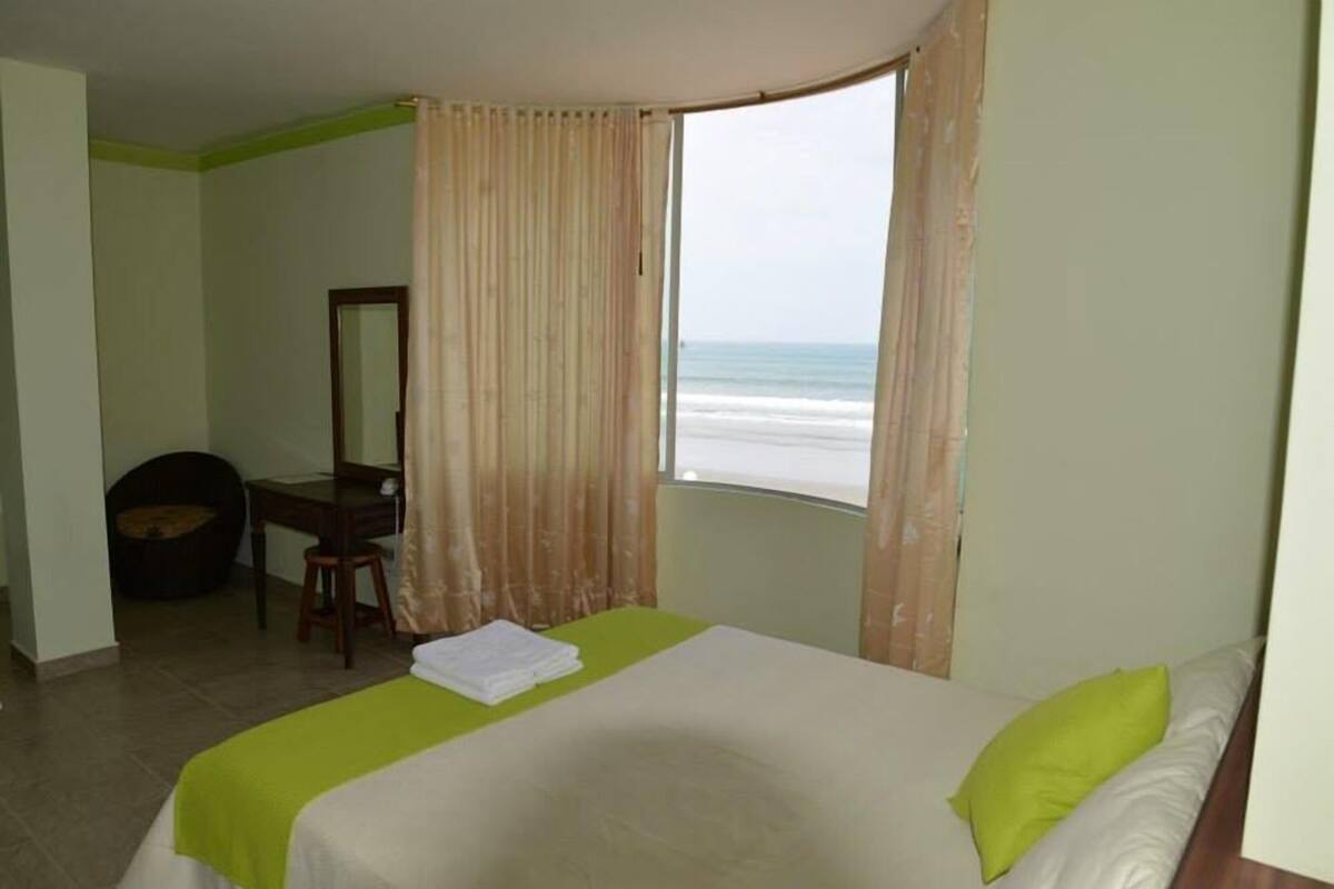 Quarto com cama, escrivaninha, cadeira e janela com vista para a praia.
