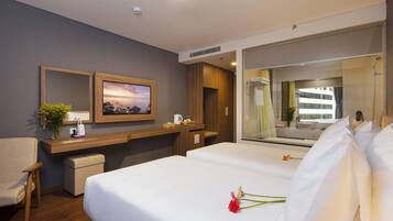 Quarto de hotel com uma cama, uma escrivaninha com um computador, uma cadeira, uma pequena mesa com um vaso de flores e um espelho grande.