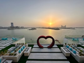 Terraço no telhado com uma escultura em formato de coração, mesas ao ar livre e vista do pôr do sol sobre a água com ilhas.
