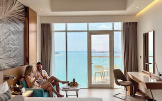 Quarto de hotel com uma janela grande, uma cama, uma escrivaninha e uma mesa de jantar com uma cesta de frutas.