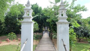 Uma ponte de madeira com pilares brancos e guarda-corpo de corda, cercada por vegetação.