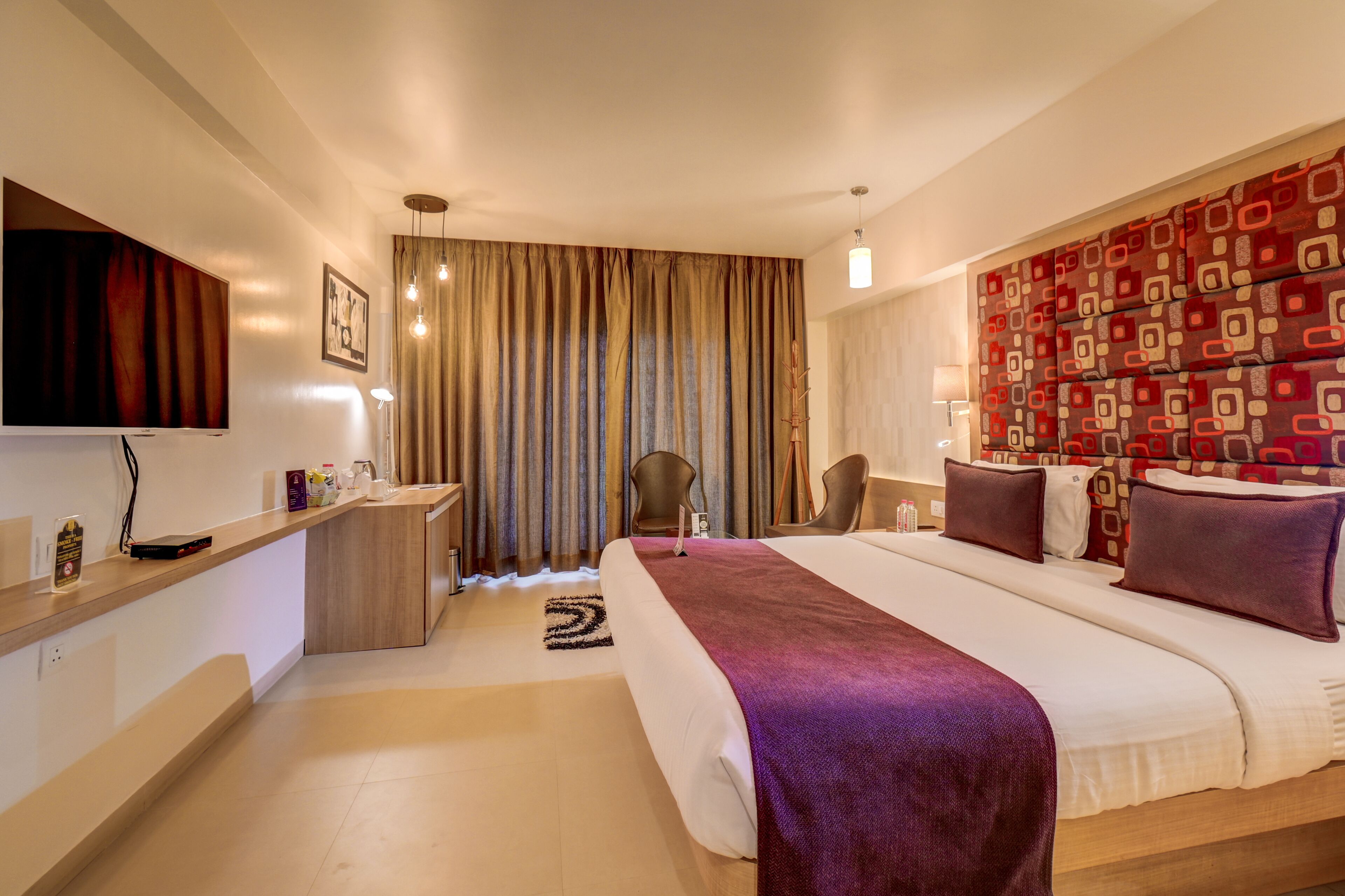 Quarto de hotel com cama, escrivaninha, cadeira, televisão e parede com padrão.