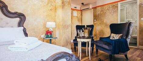 Quarto de hotel com uma cama grande, uma poltrona, uma mesinha com luminária e um minibar.
