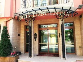 Entrada do Hotel Club Central com uma grande placa acima das portas, uma pequena árvore e um canteiro de flores à esquerda.