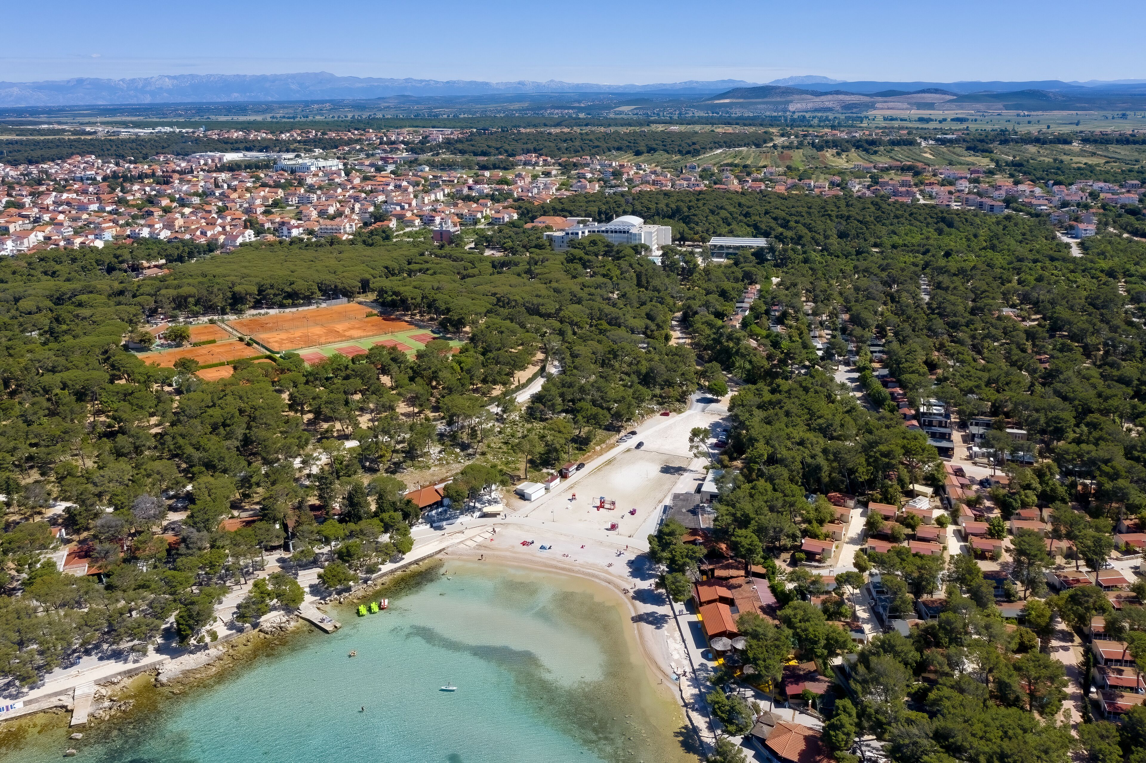 Vista aérea de uma cidade costeira com praia, áreas residenciais e um campo esportivo.