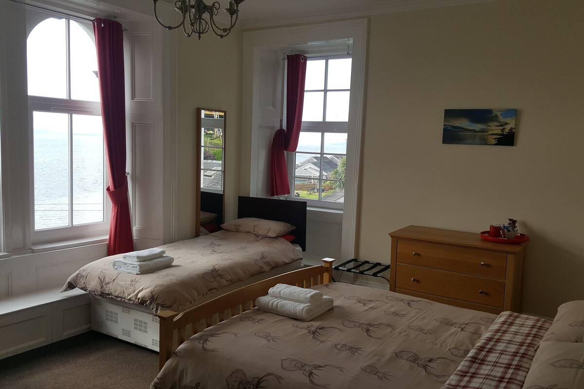 Quarto com duas camas, cómoda de madeira, espelho e janela com cortinas.