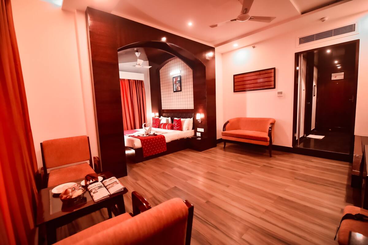 Quarto de hotel com cama, duas cadeiras, uma mesa com telefone e revistas, uma cortina vermelha e uma porta com espelho.