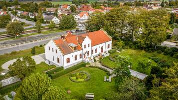 Um edifício branco e amplo com telhado de telhas vermelhas, cercado por vegetação exuberante e um gramado bem cuidado.