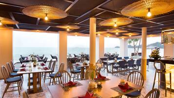 Restaurante com mesas preparadas para refeições, grandes guarda-sóis e vista para o mar.