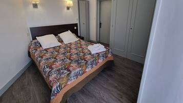 Cama bem arrumada com colcha estampada, duas luminárias na parede e um guarda-roupa com porta cinza.
