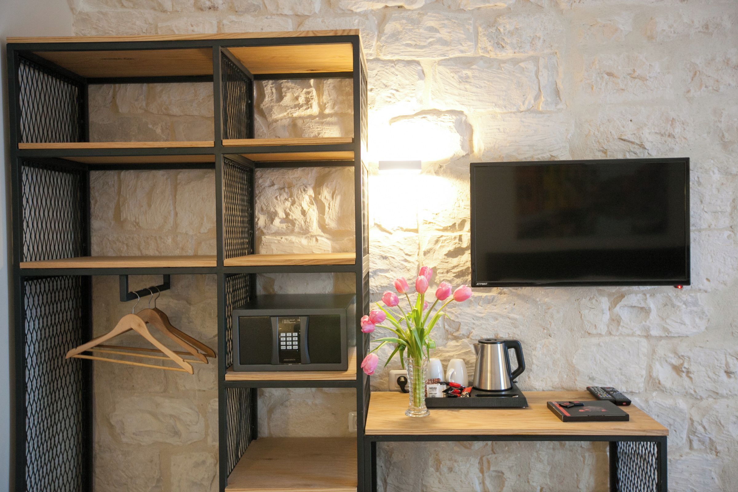 Quarto com parede de pedra, TV de tela plana presa acima de uma mesa de madeira e estante com roupas penduradas.