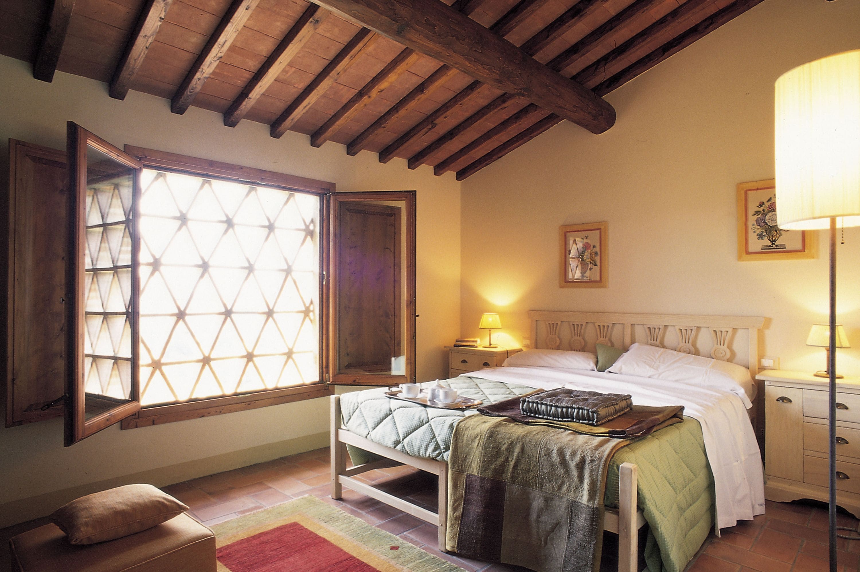 Quarto com teto de madeira, cama com colcha, mesa de cabeceira com abajur e janela com persianas de madeira.