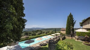 Área de piscina com espreguiçadeiras e guarda-sóis, cercada por muros de pedra e vegetação, com vista para colinas ao longe.